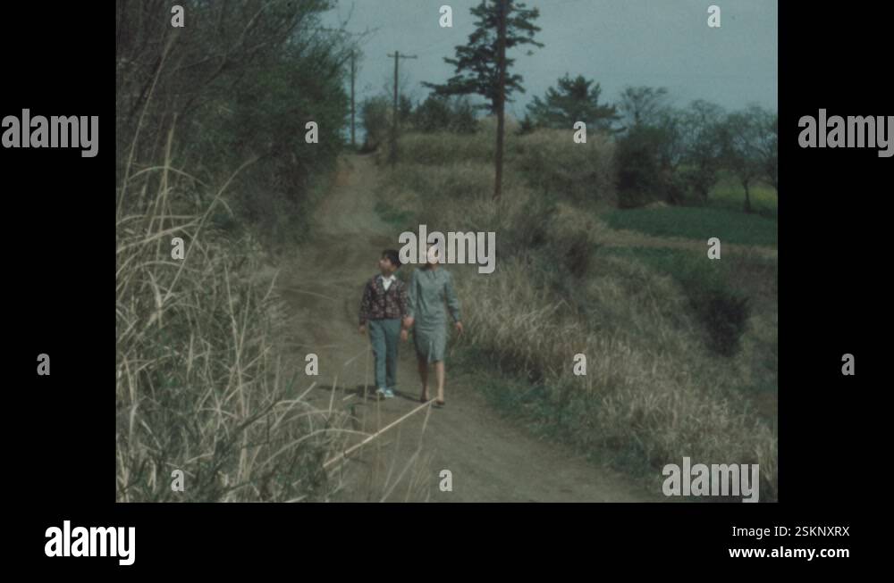 Japan 1960s: Woman and boy walk down a dirt road, woods on one side and ...