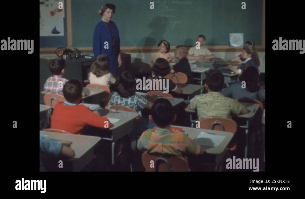 1960s: Teacher talks to a classroom of children seated at desks. A boy ...