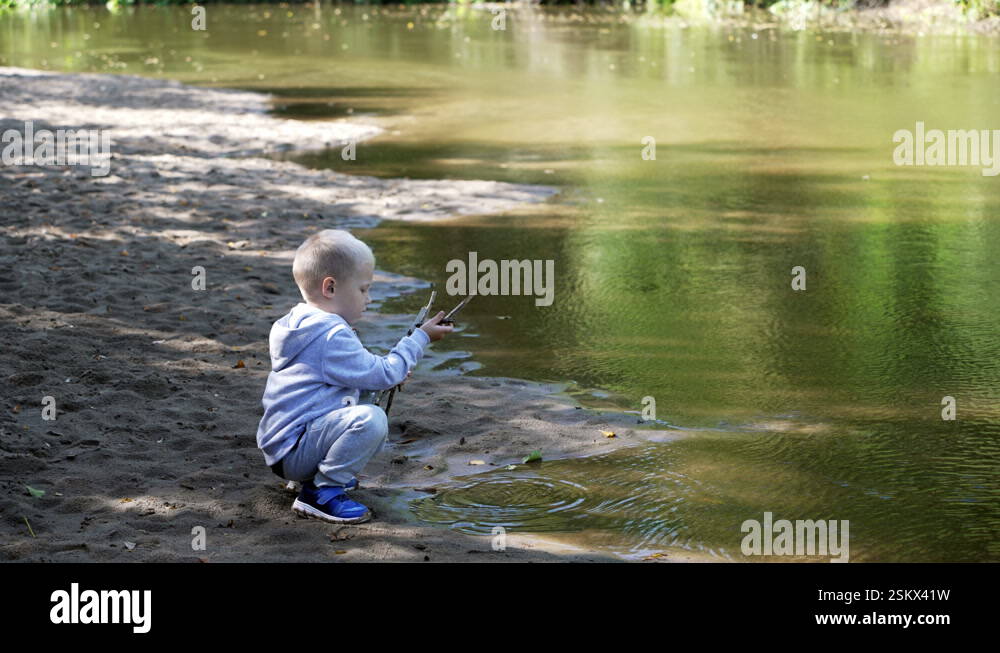 A kid boy traveler plays with sticks in the water on the river bank ...