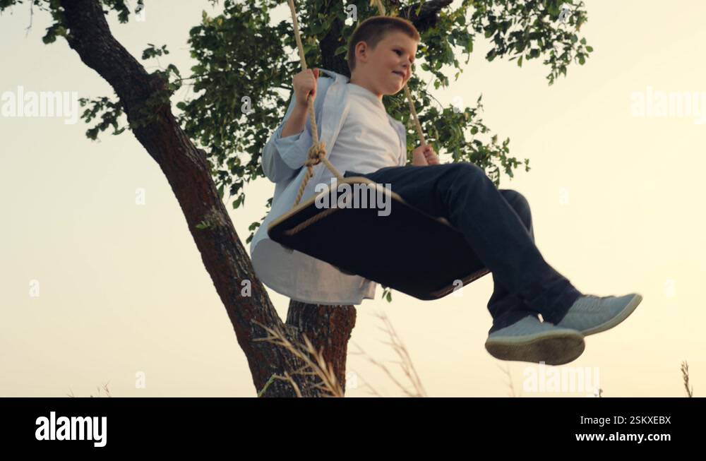 Positive preteen boy plays swings hanging on garden tree at sunset time ...