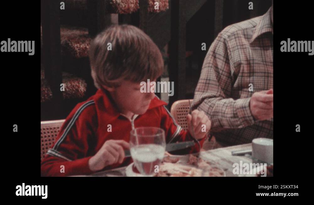 1980s: Family at dinner table. Dad cuts meat on son’s plate. Clouds in ...