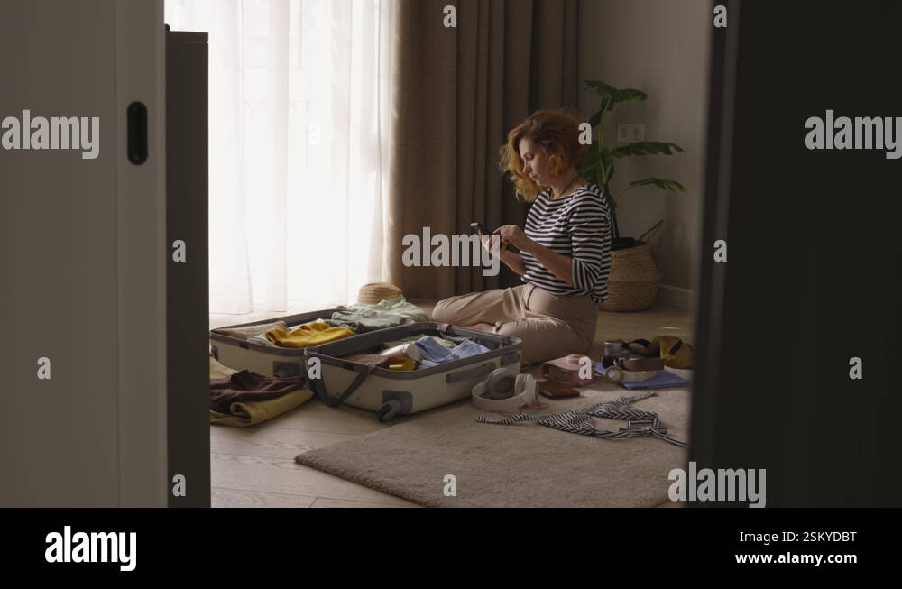 Woman packing things in suitcase, checking list on phone. Young curly ...