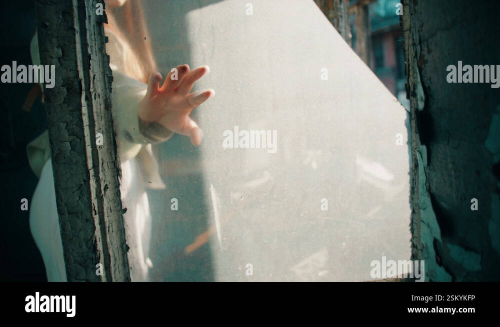 A woman's fingers touch the dusty glass of a broken window in an old ...