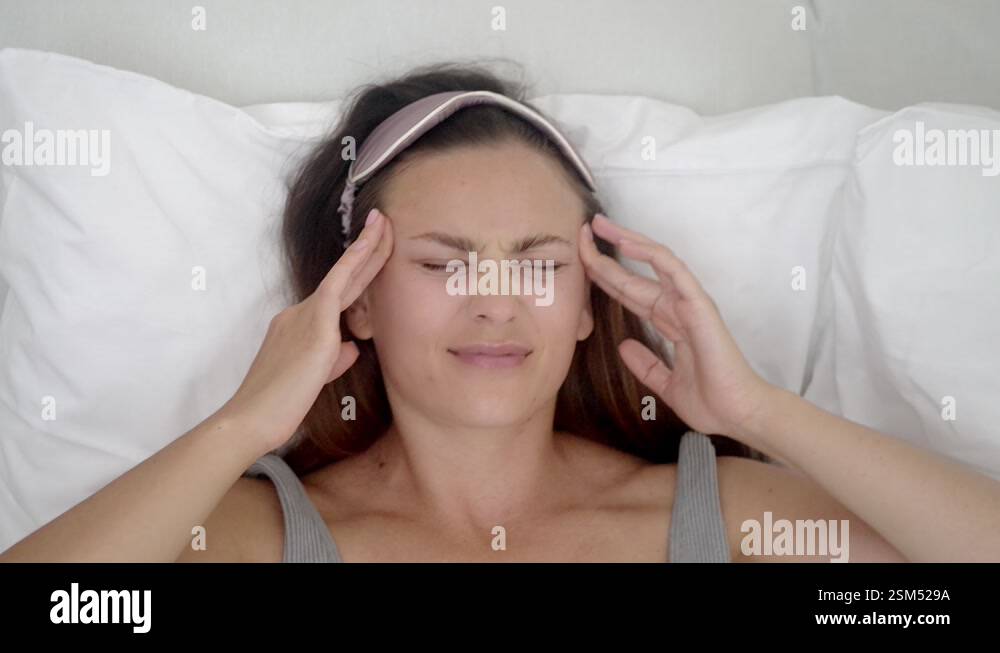 young woman, sleep mask on, frowning, massages temples, enduring a ...