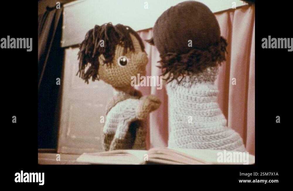 1970s: Two puppets talk to each other next to open book. Puppet touches ...