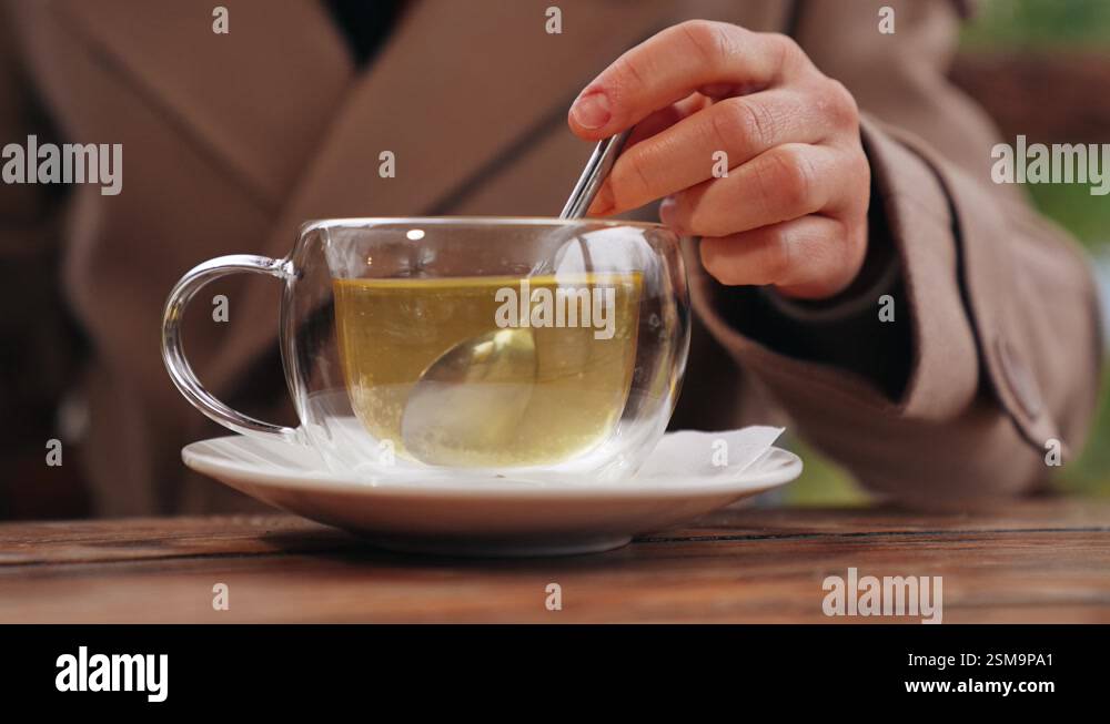 Woman stirs green tea with spoon in glass cup on table in cafe of park ...