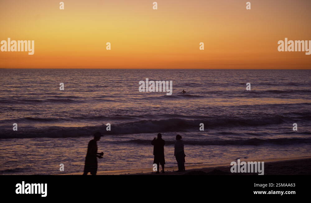 A surfer paddles out on the ocean as passers by watch the sunset on a ...