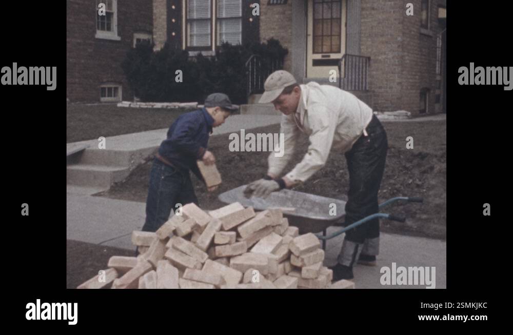 1950s: Man and boy load bricks into wheelbarrow. Boy feels arm muscle ...