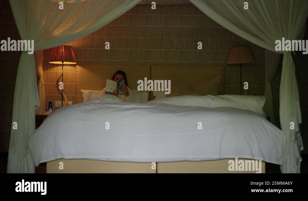 Women lying in the bed of a luxury hotel reading a book with a bedside ...