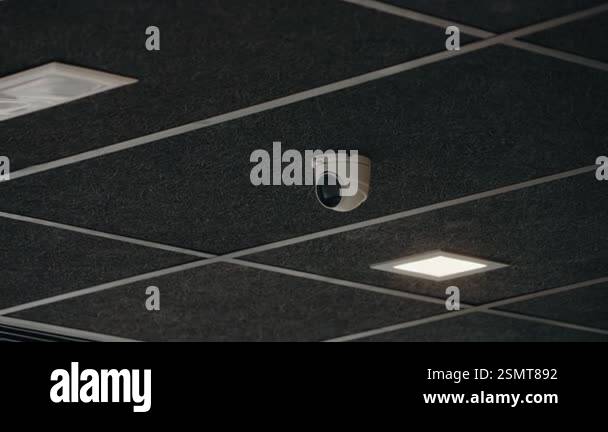 A white security camera hangs from a dark, textured ceiling. The ...