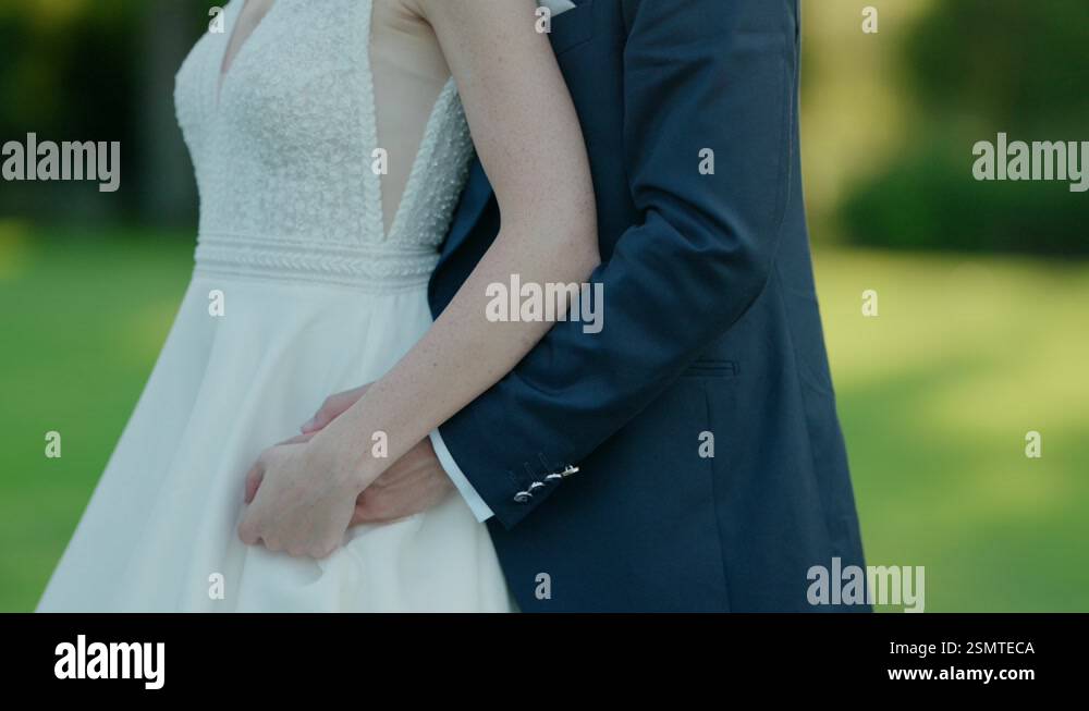 bride and groom embrace, his arm around her waist, sunlight casts soft ...