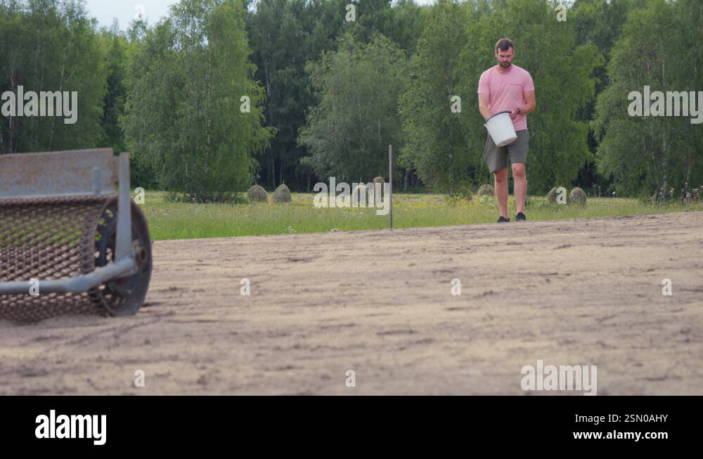 A man sowing grass on a field near a forest in the summer with a tool ...