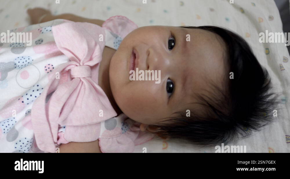 Baby girl in a pink bow gazes at the camera while wriggling on her bed ...