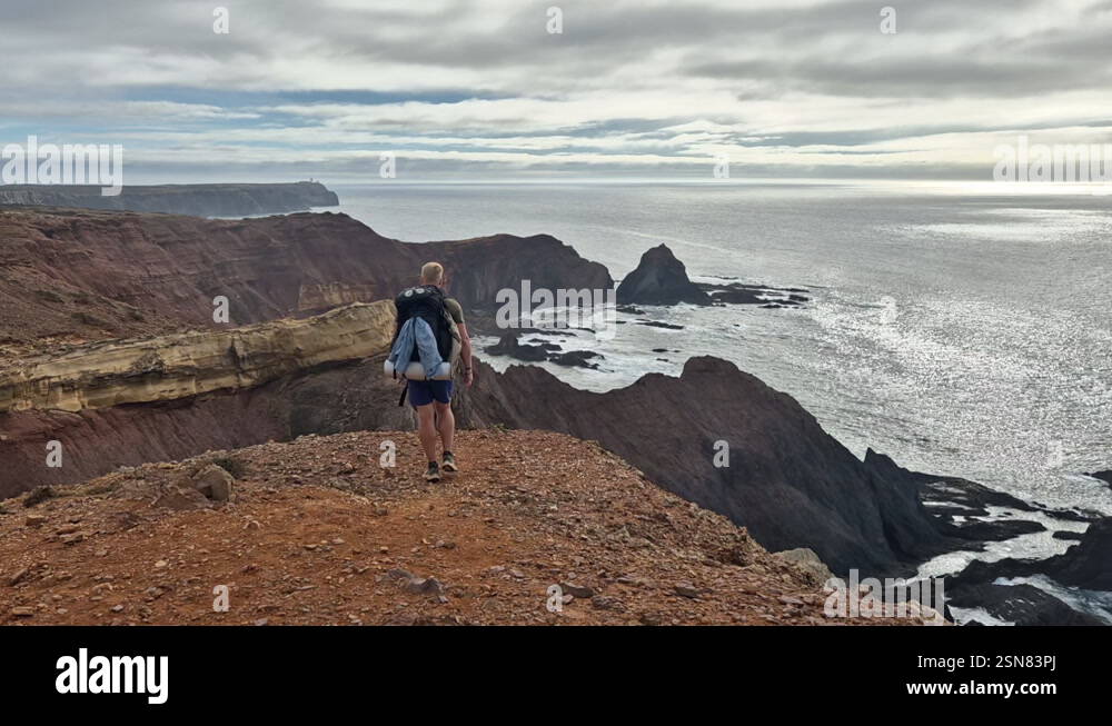 Male backpacker walks to edge of tall ocean cliff to enjoy the view ...