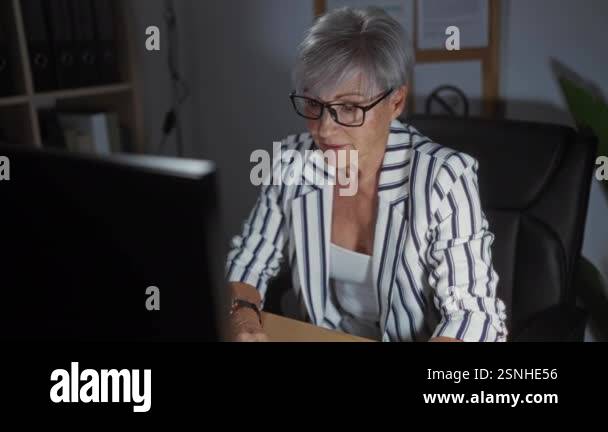 Middle-aged woman with grey hair and glasses working in an office at ...