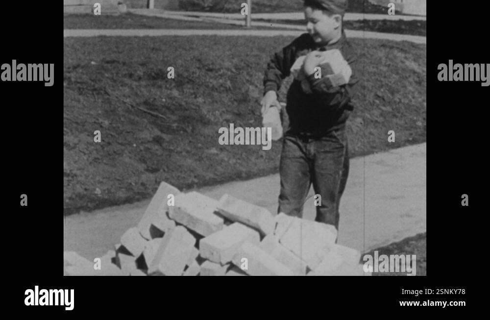 1950s: Young boy gathers bricks from pile. Boy carries bricks past ...