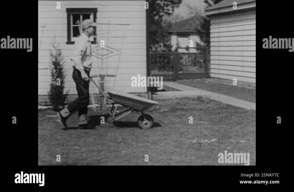 1950s: Man pushes wheelbarrow through backyard as boy points. Steam ...
