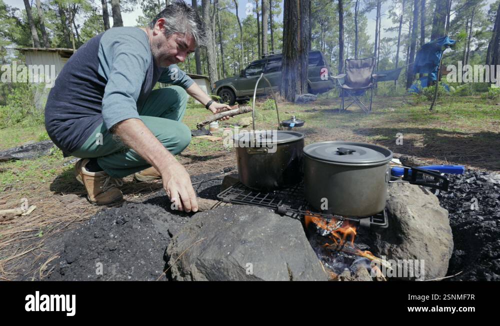 Man adding wood to a campfire cooking in a forest with rustic setup and ...