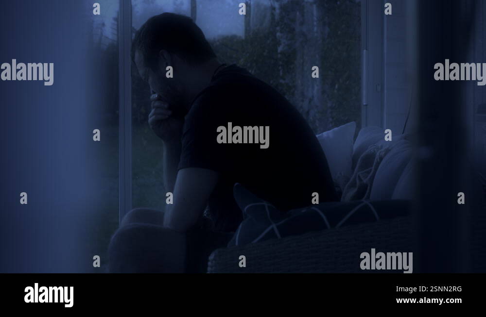 Man sitting by a window at night in deep thought with dim lighting and ...