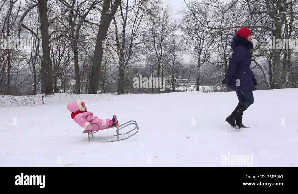 Mother pull baby girl on sledge along snow in winter resort. Handheld ...