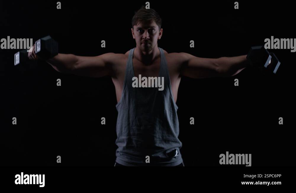 4K Fit young man with muscular physique lifting weights against black ...