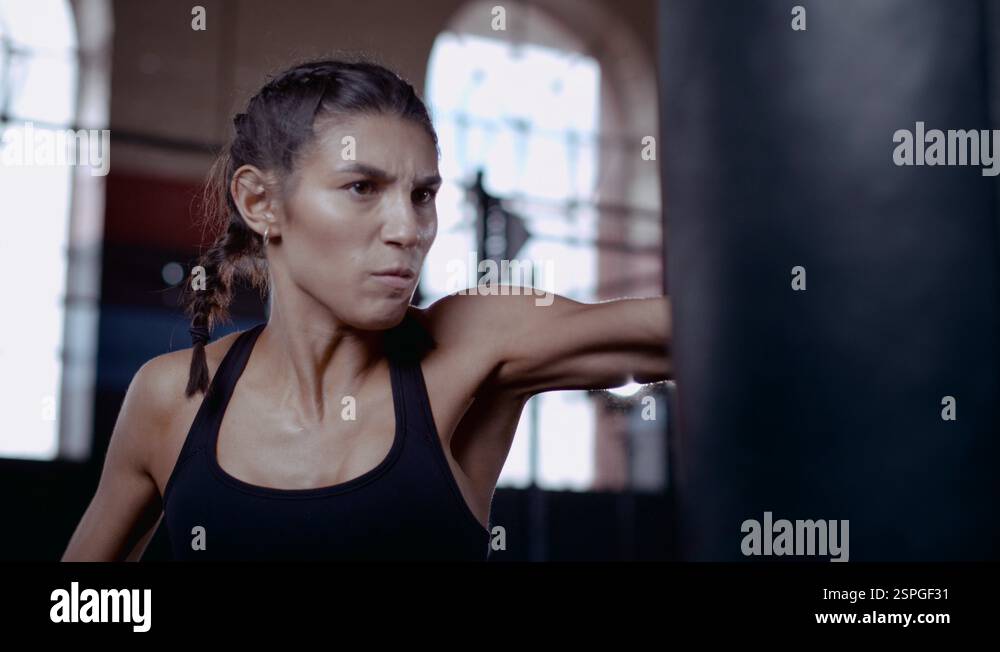 4K Powerful female boxer training with punch bag in urban gym. Super ...