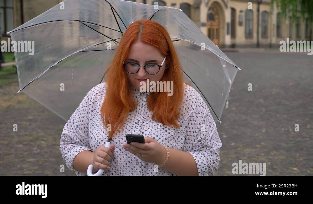 Fat ginger girl with glasses is typing message on smartphone in park in rainy Stock Video ...