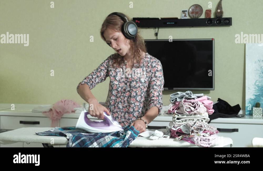 woman ironing the mountain of laundry at home in the kitchen listening ...