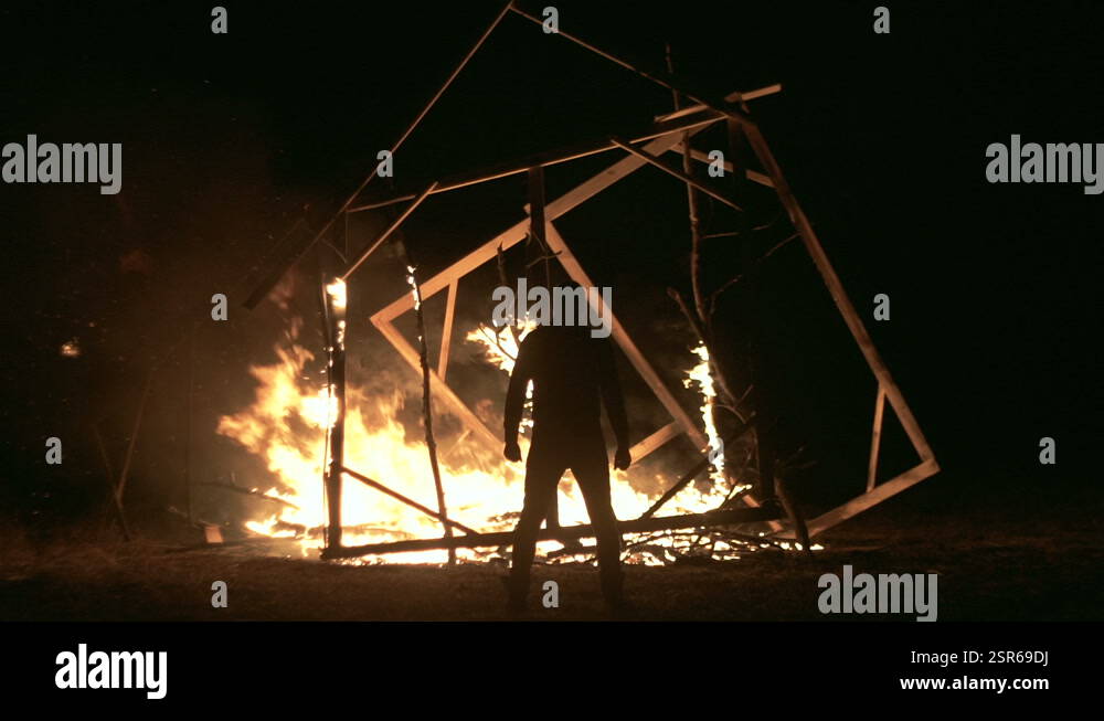 A man stands at a burning house, a man against a burning house at night ...