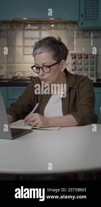 A mature woman writes with her left hand while working on a laptop at ...