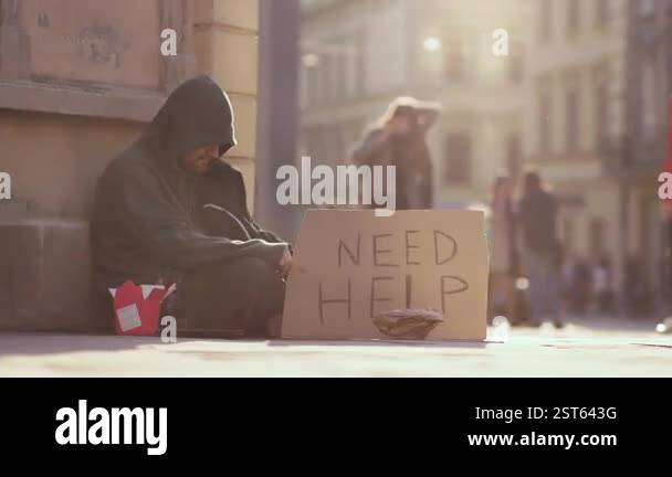 A vulnerable figure sits on the street, seeking help with a handwritten ...