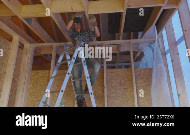 A construction worker is positioned on a ladder to install wooden beams ...