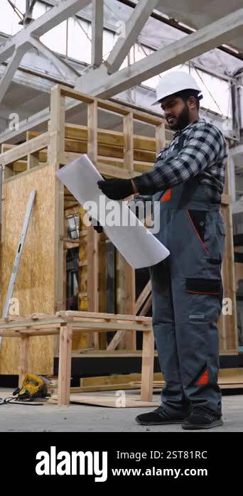 An Indian engineer wearing a hard hat and gloves examines blueprints while overseeing the ...