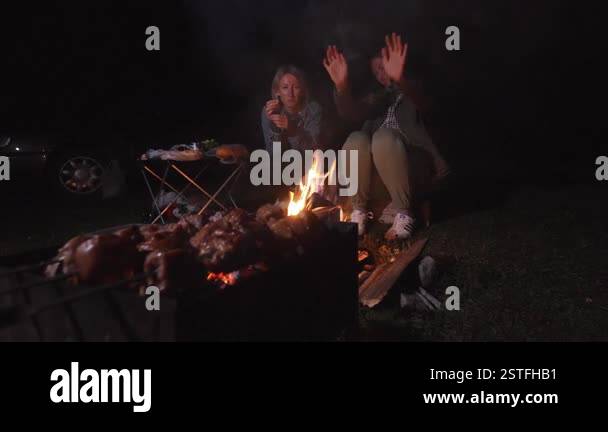 Two girlfriends of girls enjoying warmth and food on grill by fire ...