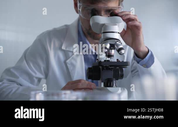 Male scientist wearing lab coat and protective glasses using microscope ...