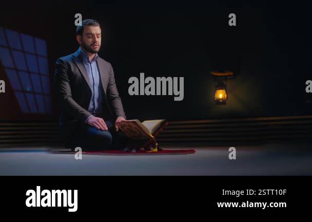 Muslim man in formal attire sits on a prayer mat, reciting from an open ...