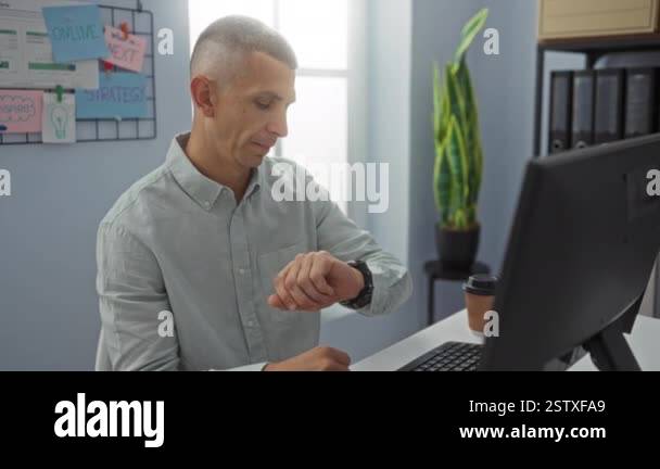 Man working in an office looking at watch while sitting at a desk with ...