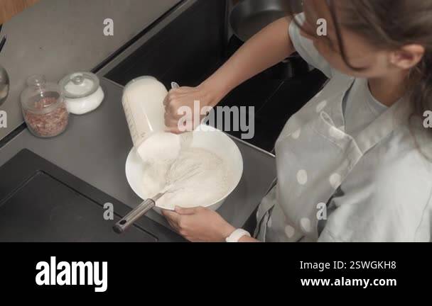 A woman pours flour into liquid batter using a measuring cup and mixes ...