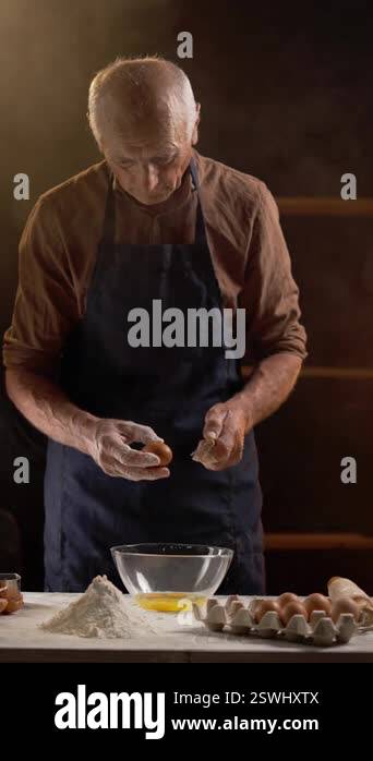 Elderly baker in an apron cracking an egg into a transparent bowl ...