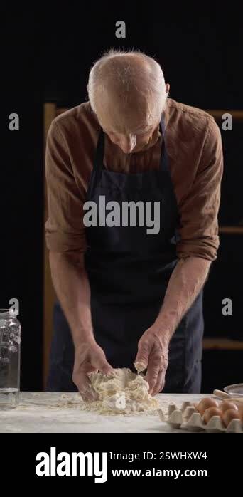 Elderly baker in an apron kneading dough by hand on a floured surface ...