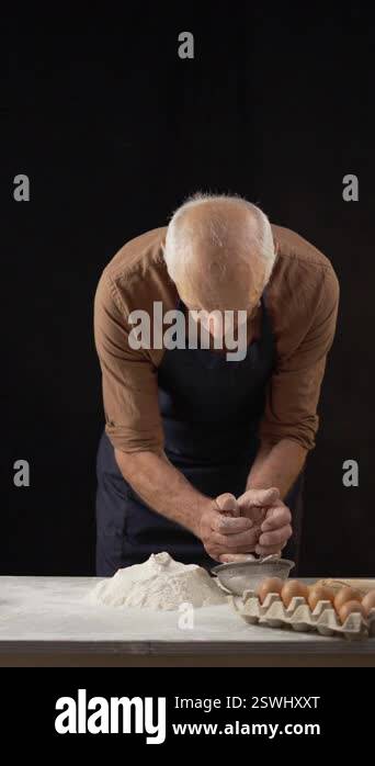 Elderly baker in an apron pouring flour into a regular sifter over a ...