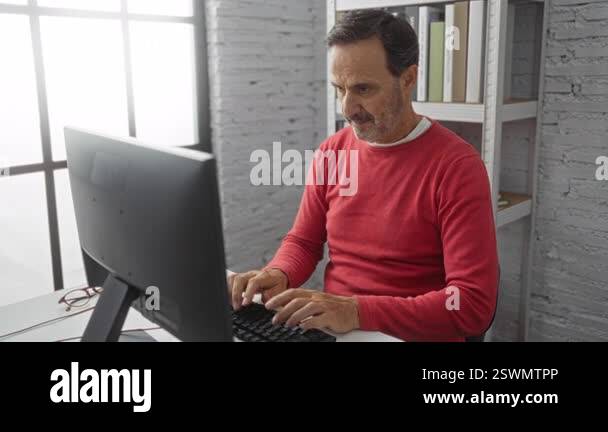 Man working on computer in a modern office, wearing casual red sweater ...