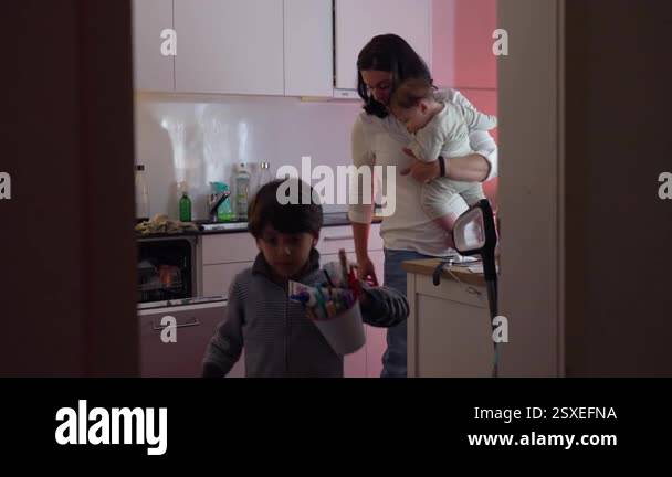 Mother multitasking in kitchen holding baby while older child carries a ...