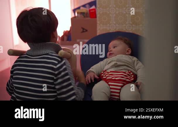 Young boy sitting on floor holding chair near baby on bouncer, baby watching sibling attentively ...