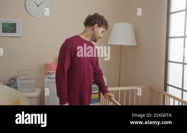 Young man in a bedroom stands by a baby cradle with a gentle smile ...