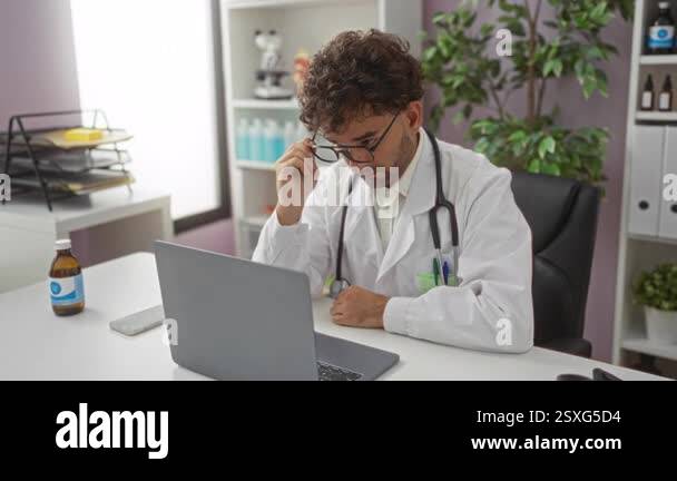 Hispanic male doctor with glasses focuses intently on a laptop in a ...