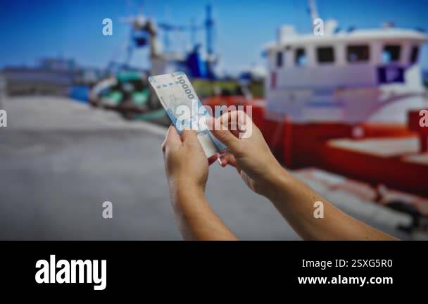 Hands holding chilean pesos outdoors in a seaside port with boats, depicting business and ...