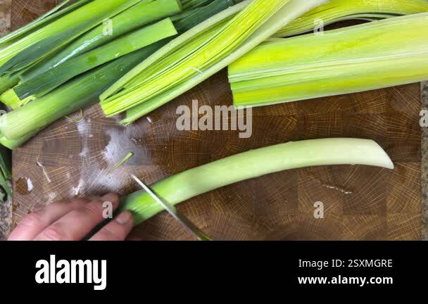 A cook cuts leek stems with a kitchen knife on a wooden cutting board ...