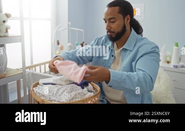 Man in bedroom organizing baby clothes in a basket with a crib nearby ...