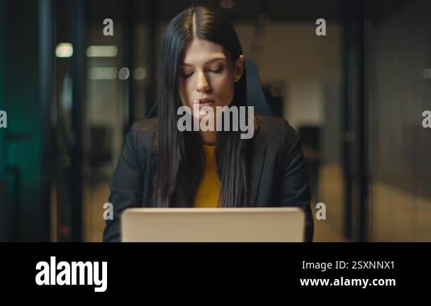 Stressed businesswoman in a dark office, deeply focused on her laptop ...
